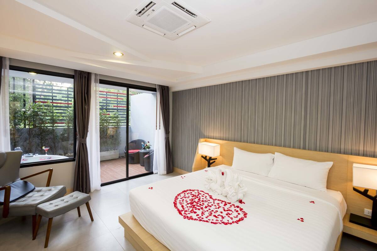Quarto de hotel com uma cama grande, uma mesa e uma cadeira. Possui uma varanda com vista para o verde e uma mesa com cadeiras.