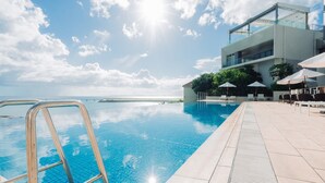 Uma piscina com escada metálica, área de estar com cadeiras e guarda-sóis, e um edifício moderno com fachada de vidro.