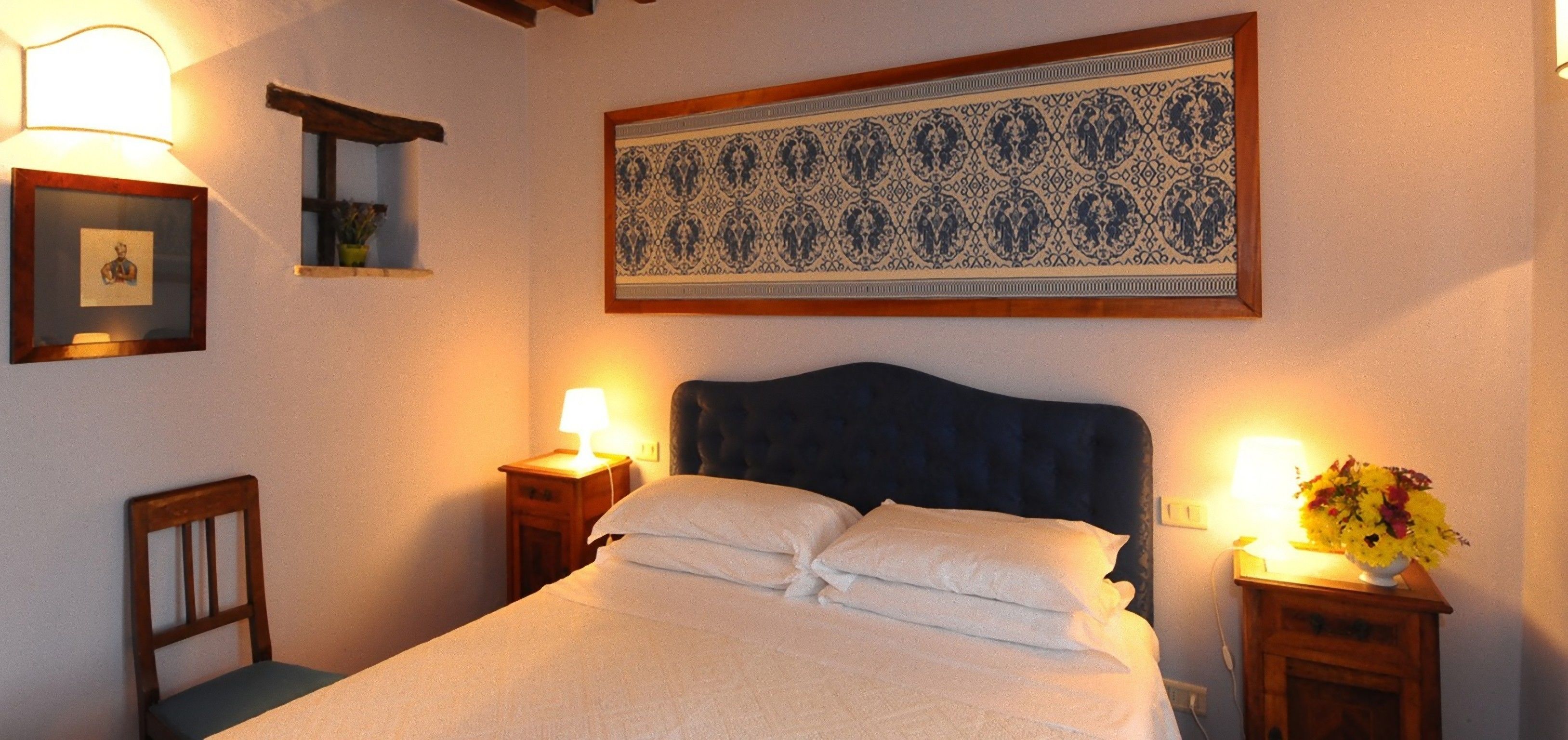 Cama bem arrumada com lençóis brancos, duas luminárias laterais, cabeceira de madeira, painel decorativo na parede, uma mesa pequena com um vaso de flores e uma cadeira.
