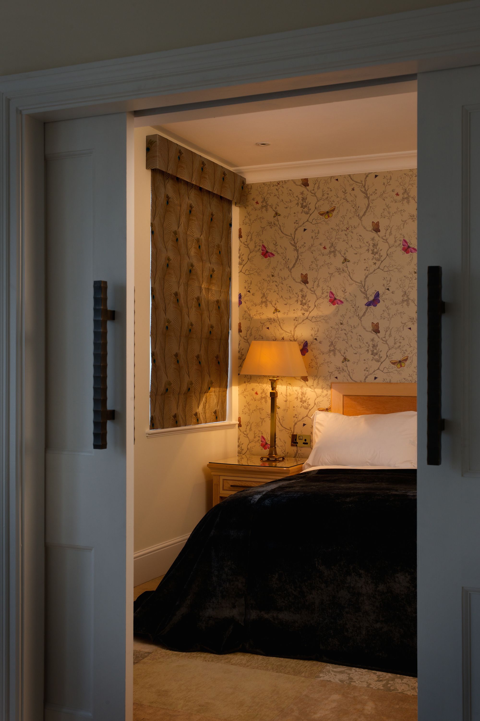 Quarto de hotel com cama, mesa de cabeceira, abajur e janela com cortinas.