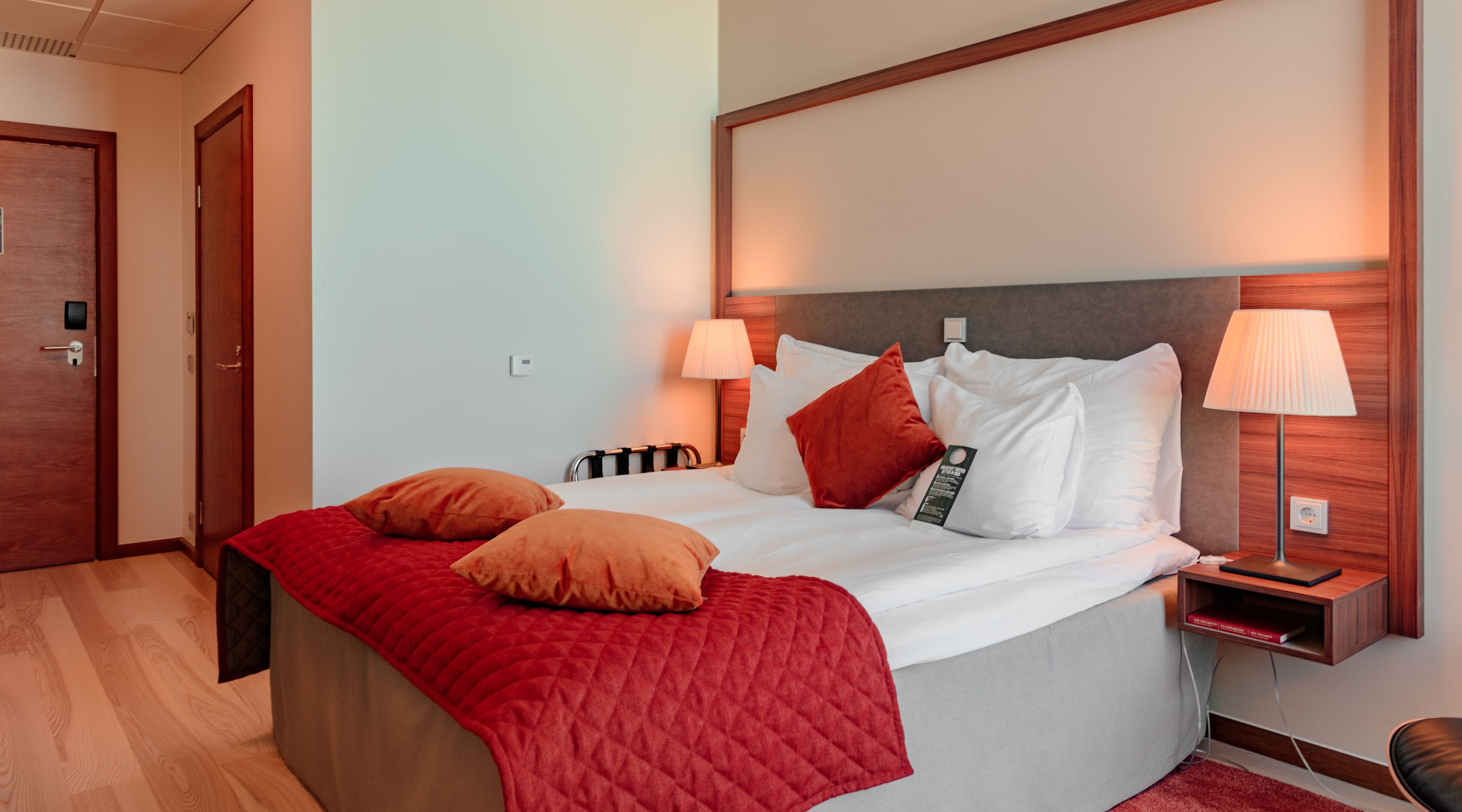 Quarto de hotel com cama, duas luminárias de cabeceira, mesa de cabeceira e manta vermelha.