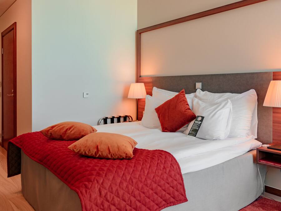 Quarto de hotel com cama, duas luminárias de cabeceira, mesa de cabeceira e manta vermelha.