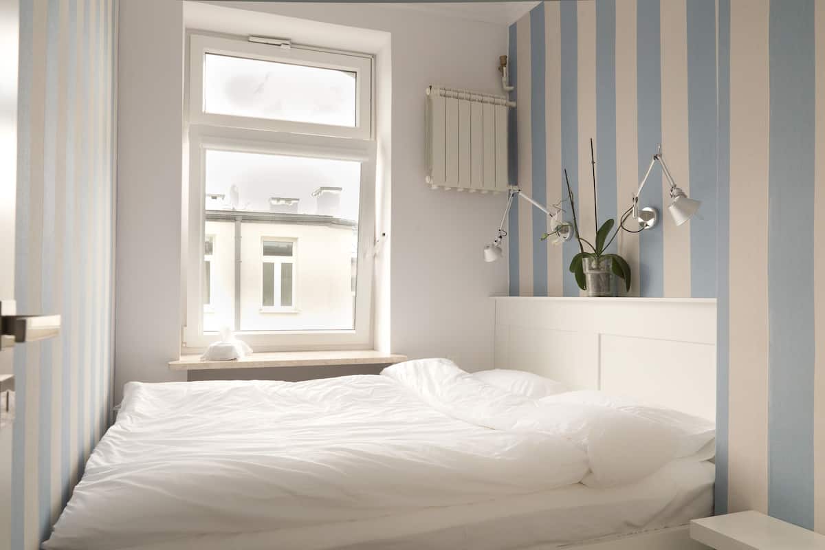 Quarto com cama, janela, radiador e uma planta sobre uma mesinha de cabeceira.