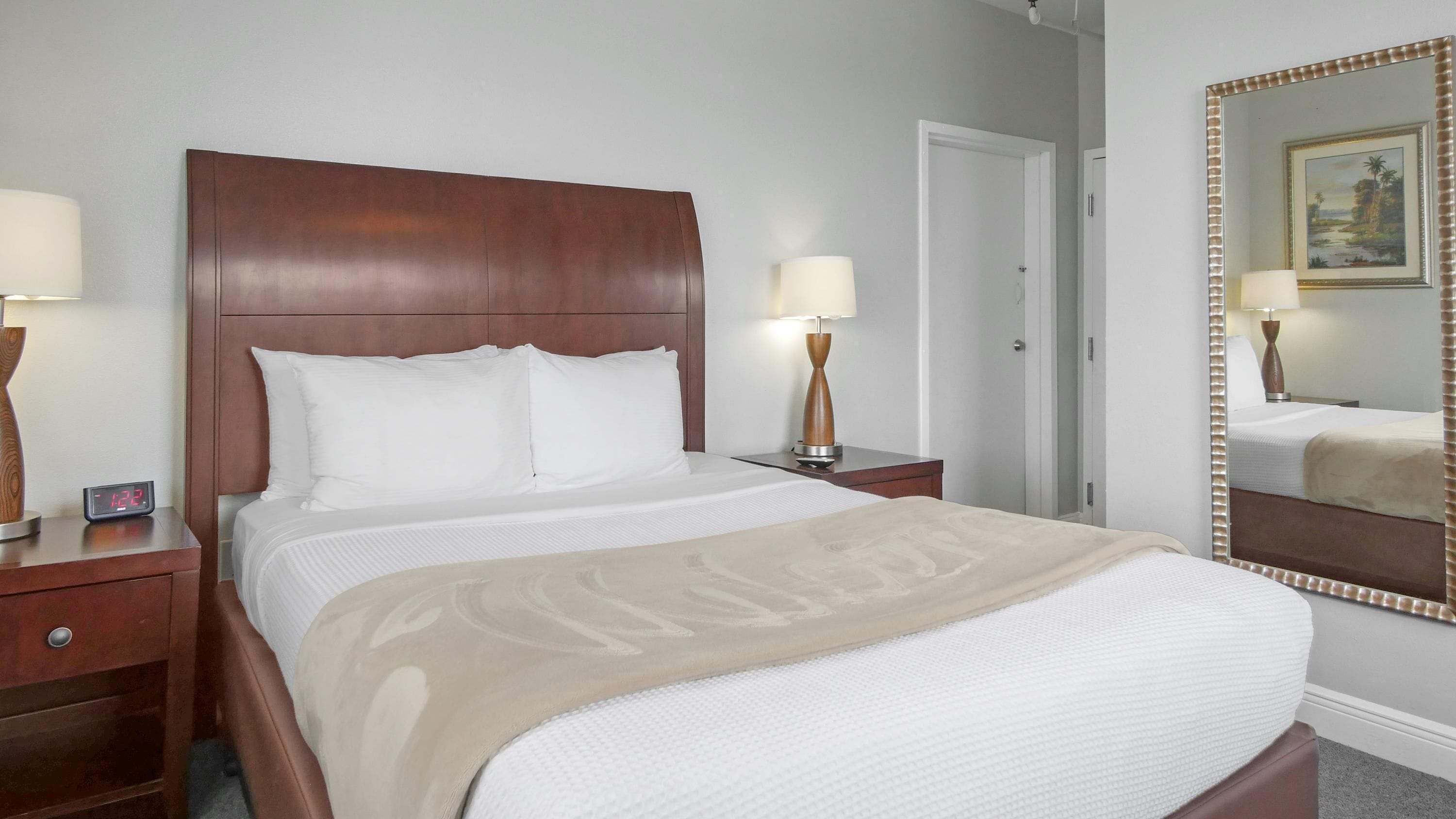 Quarto de hotel com uma cama grande, cabeceira de madeira, duas mesas de cabeceira com luminárias, um criado-mudo e um espelho com moldura decorativa, além de uma porta.