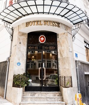 Entrada do Hotel Suisse, com uma placa destacada, símbolo de uma cruz vermelha e uma porta de vidro.