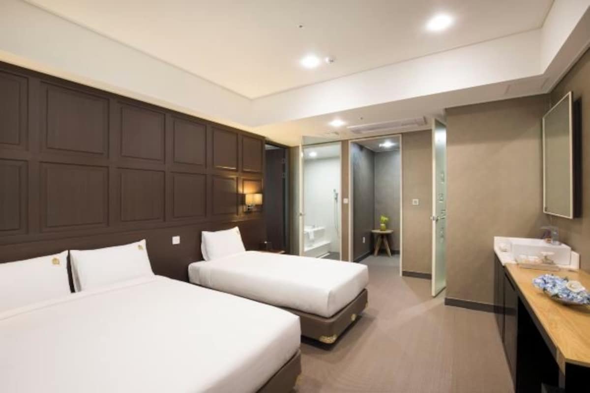 Quarto de hotel com duas camas, uma escrivaninha com espelho e banheiro visível através de uma porta de vidro.