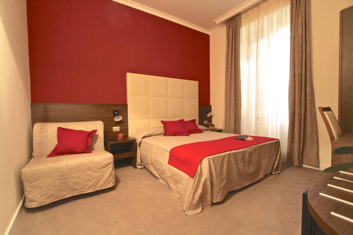 Quarto de hotel com duas camas, uma parede de destaque vermelha e uma janela grande com cortinas.