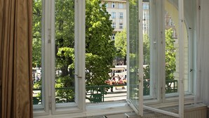 Quarto com uma janela grande que dá para um parque com árvores e um edifício ao fundo.
