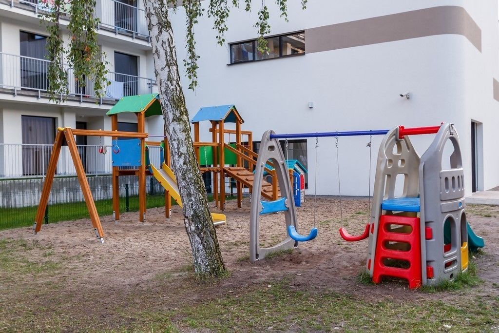 Um parquinho com diversas estruturas de recreação, incluindo um escorrega e balanços, cercado por um edifício com varandas.