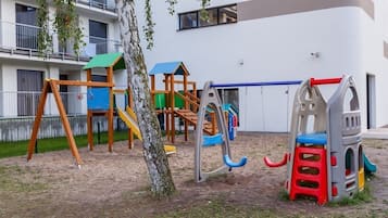 Um parquinho com diversas estruturas de recreação, incluindo um escorrega e balanços, cercado por um edifício com varandas.