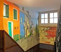 Um mural de uma rua colorida em uma parede de escadaria, mostrando um edifício azul com uma porta verde e um edifício amarelo com fachada de tijolos.