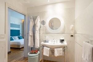 Um banheiro moderno com uma pia grande, espelho e porta-toalhas. Uma cama é visível através de uma porta aberta ao fundo.