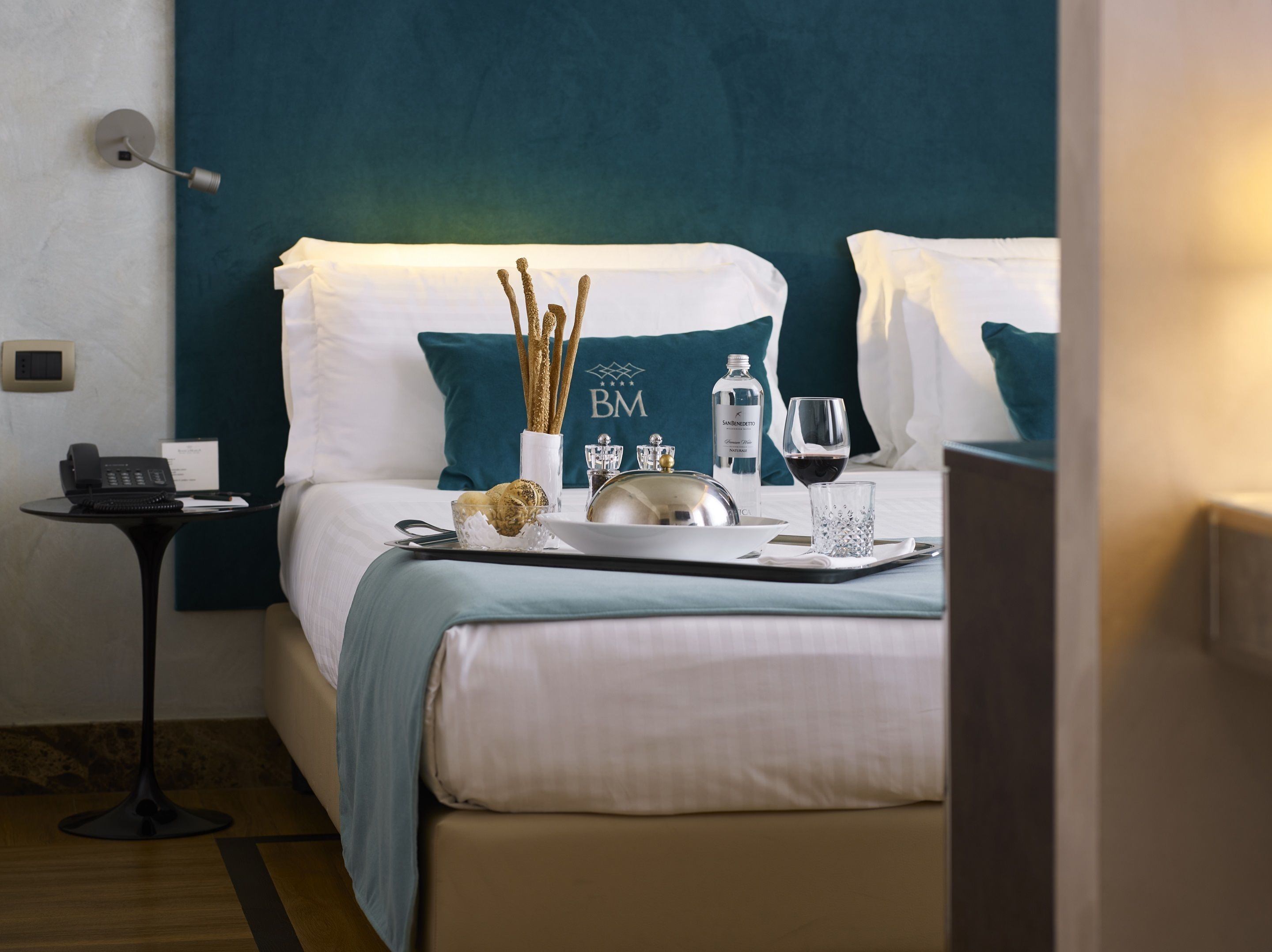 Quarto de hotel bem arrumado com cama, mesa de cabeceira com telefone e um aviso, bandeja com um copo de vinho, garrafa e objetos decorativos.