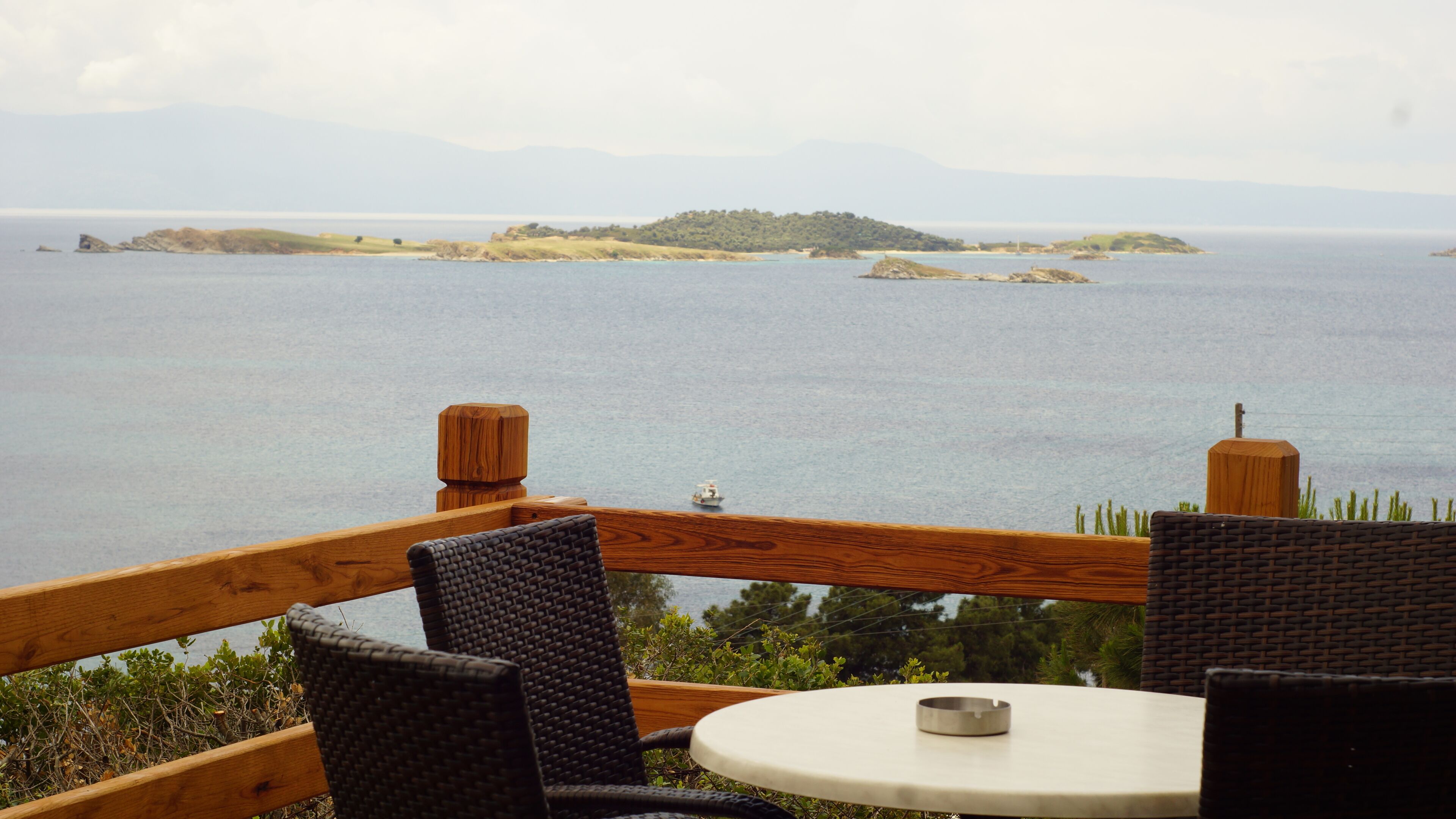 Um deck de madeira com cadeiras de vime e uma mesa, com vista para um mar calmo e ilhas ao longe.