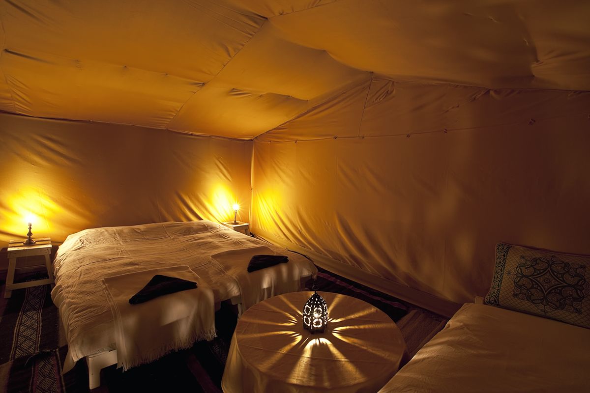 Quarto com tenda, duas camas, uma mesa pequena com abajur e luminária decorativa pendurada.