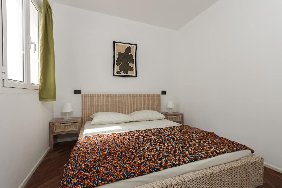 Quarto com cama, mesa de cabeceira, abajur, quadro na parede e janela com cortina.
