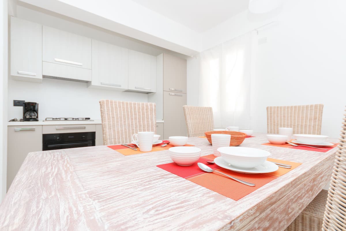 Cozinha moderna com armários brancos, cafeteira e área de refeições com mesa posta para quatro pessoas.