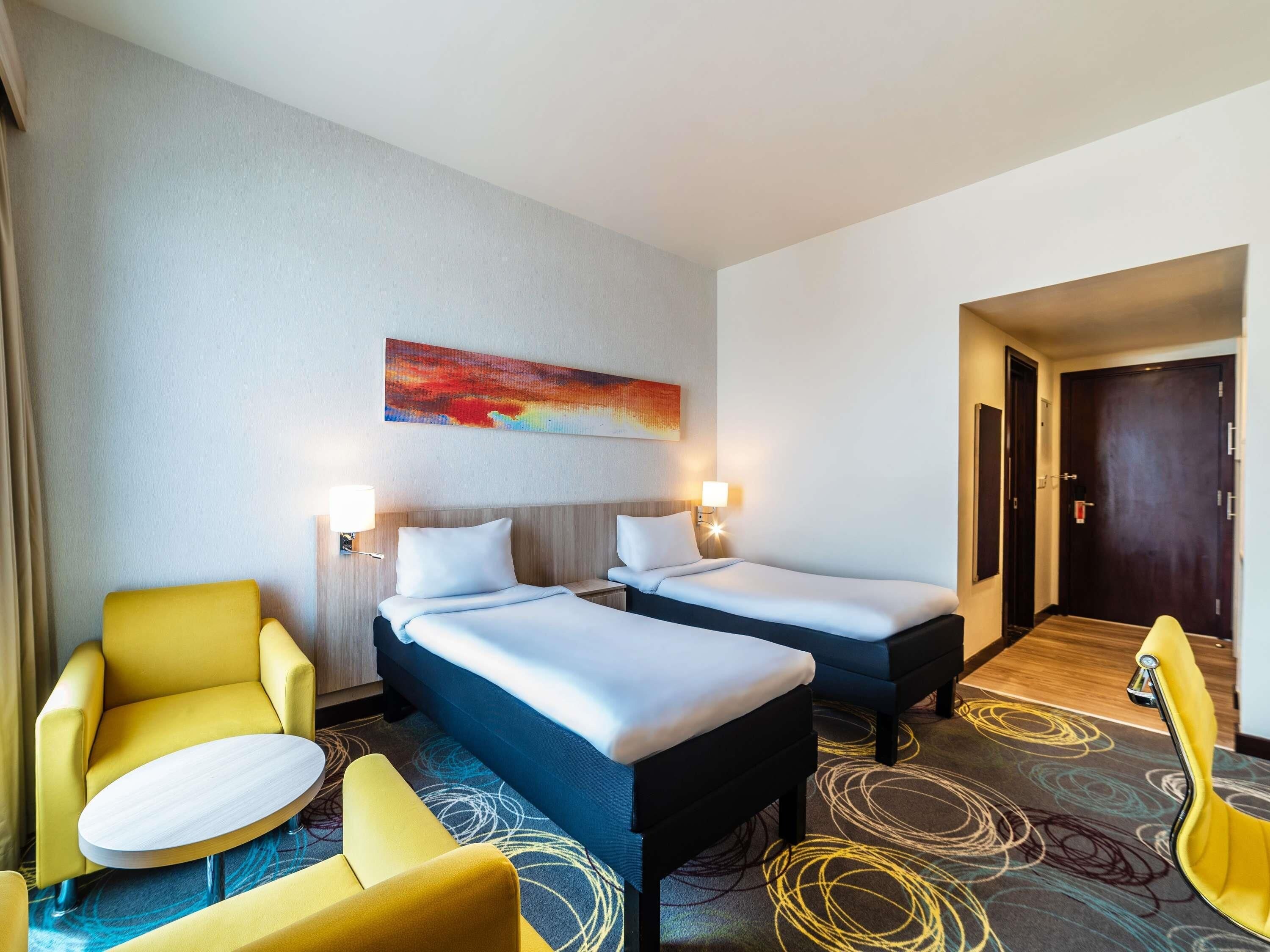 Quarto de hotel com duas camas, uma poltrona amarela, uma mesinha redonda e um quadro na parede.