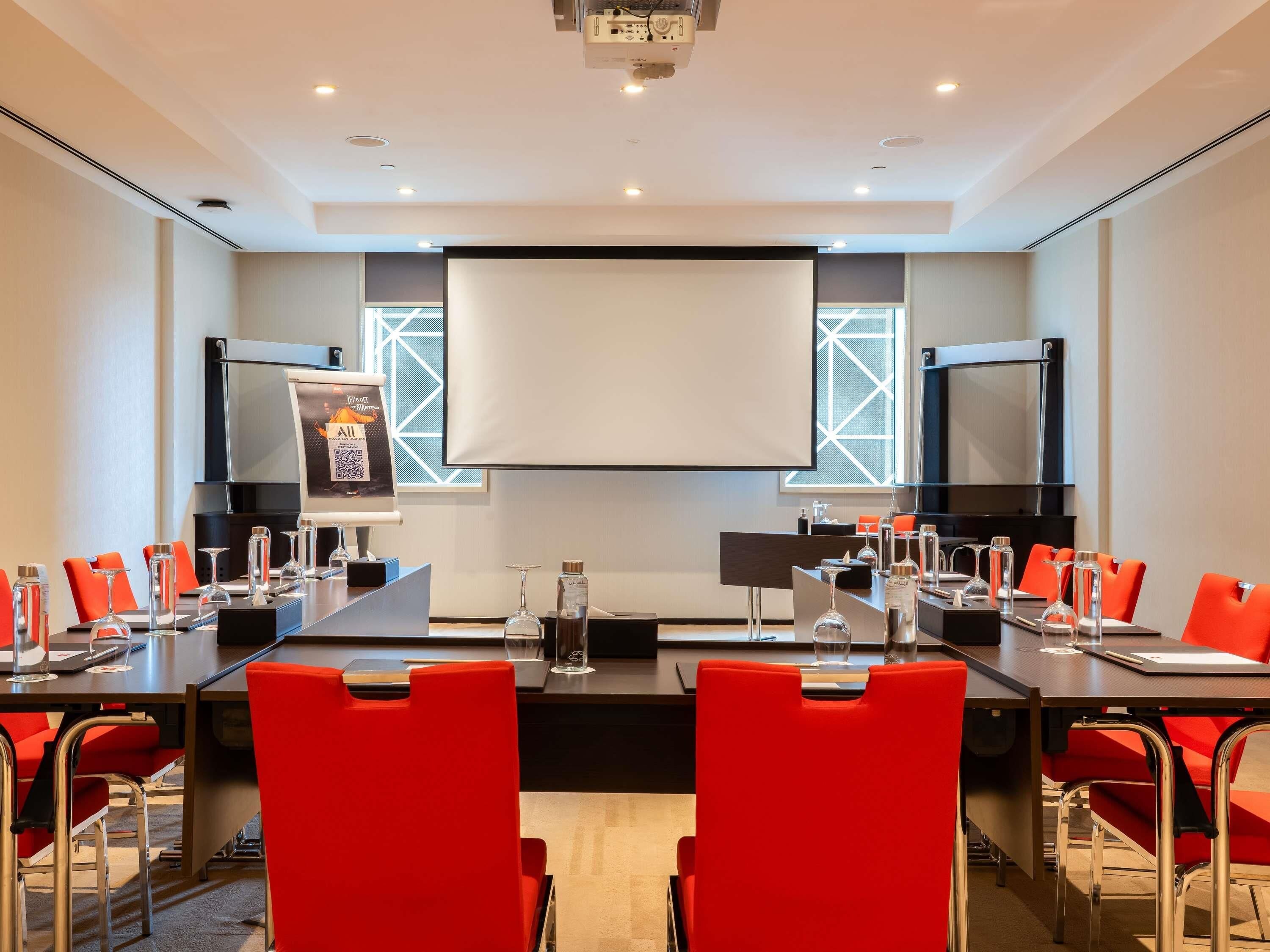 Sala de conferências com uma tela grande, várias mesas e cadeiras vermelhas.