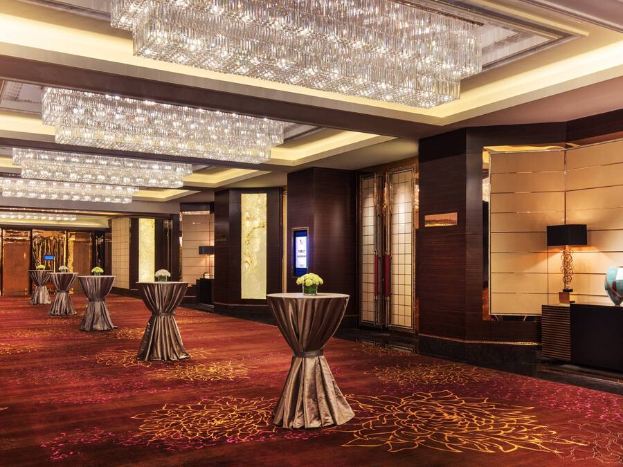 Um saguão de hotel com tapete vermelho, várias mesas redondas com toalhas e um grande candelabro.