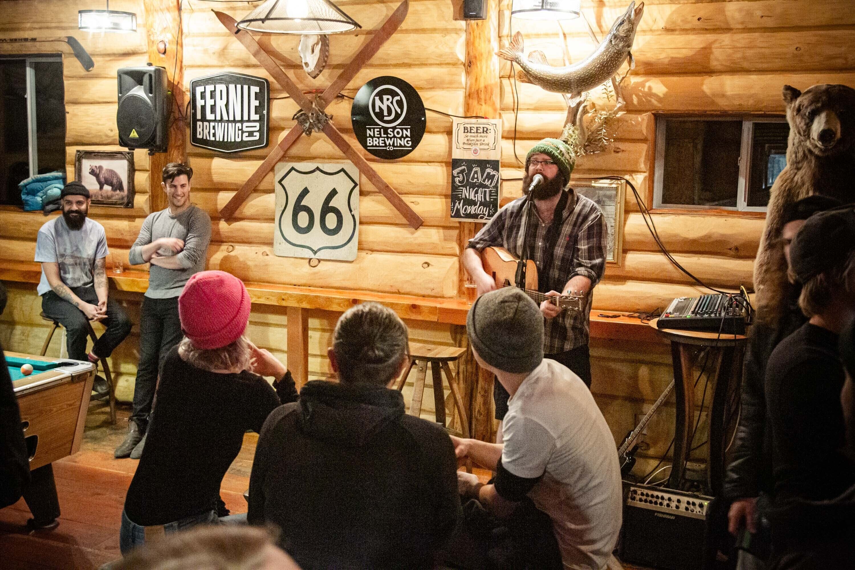 Interior rústico de madeira com palco para música ao vivo, mesa de bilhar e diversos cartazes, incluindo 'Fernie Brewing' e 'Nelson Brewing'.