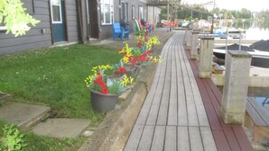 Um cais de madeira leva a uma marina com barcos, uma área gramada com vasos de plantas e um edifício com fachada escura e uma placa com a inscrição "LGS LANCING".