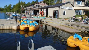 Uma marina com pedalinhos amarelos e azuis atracados, um edifício com a inscrição 'MARINA' e um letreiro para aluguel de barcos, e um cais com barcos e um pequeno píer.