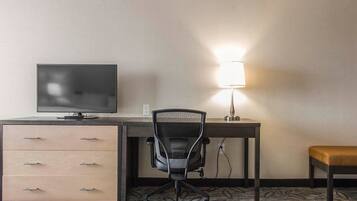 Quarto de hotel com escrivaninha, televisão, luminária, cadeira e um pequeno banco.