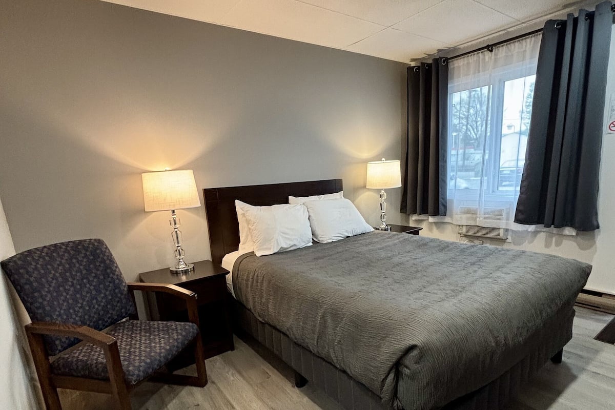 Quarto de hotel com cama grande, duas luminárias de cabeceira, uma cadeira e janela com cortinas.
