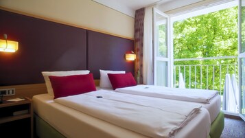 Quarto de hotel com duas camas, uma mesa de cabeceira, uma luminária e janela com vista para árvores.