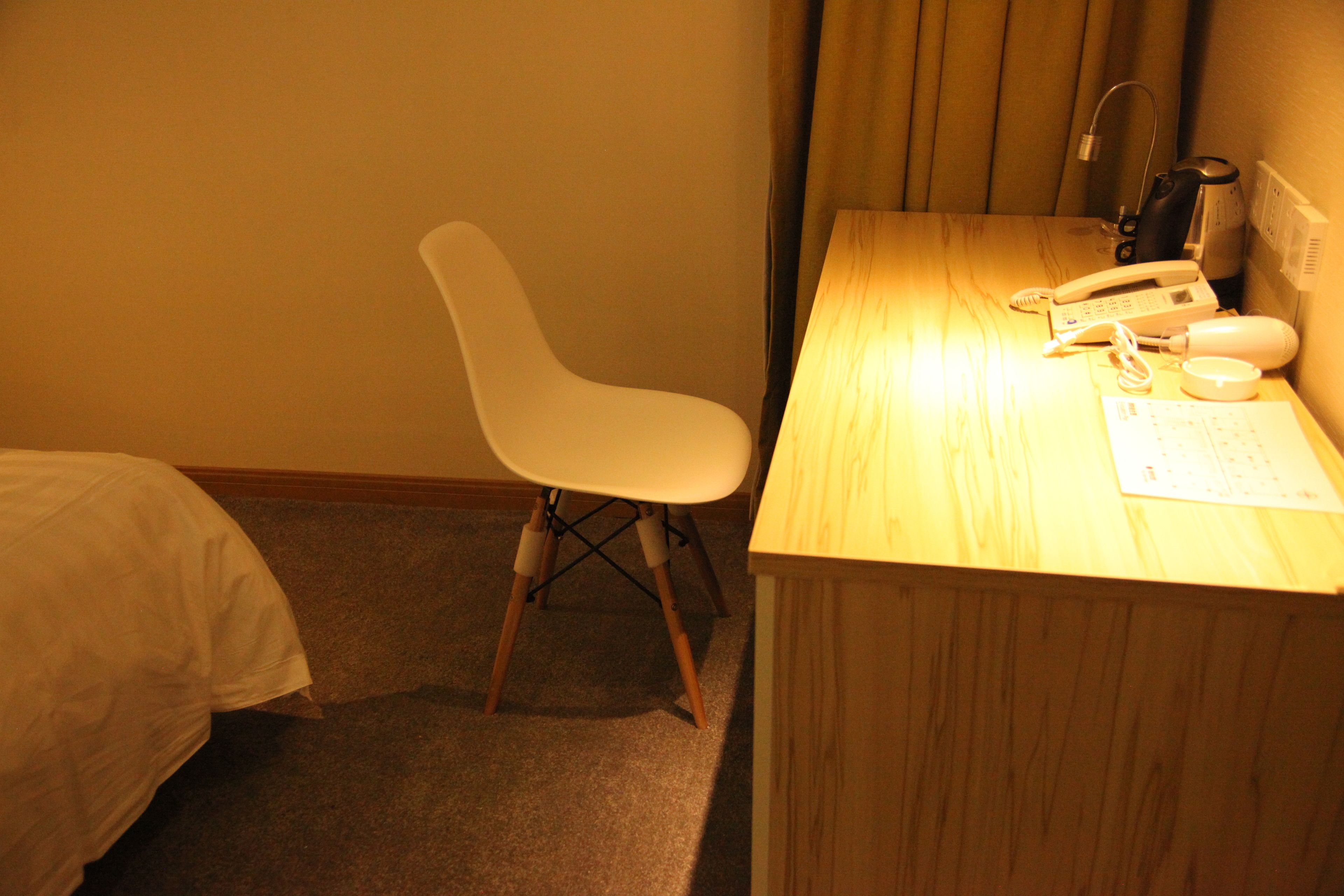 Quarto de hotel com uma secretária de madeira, uma cadeira branca, um telefone e uma luminária.