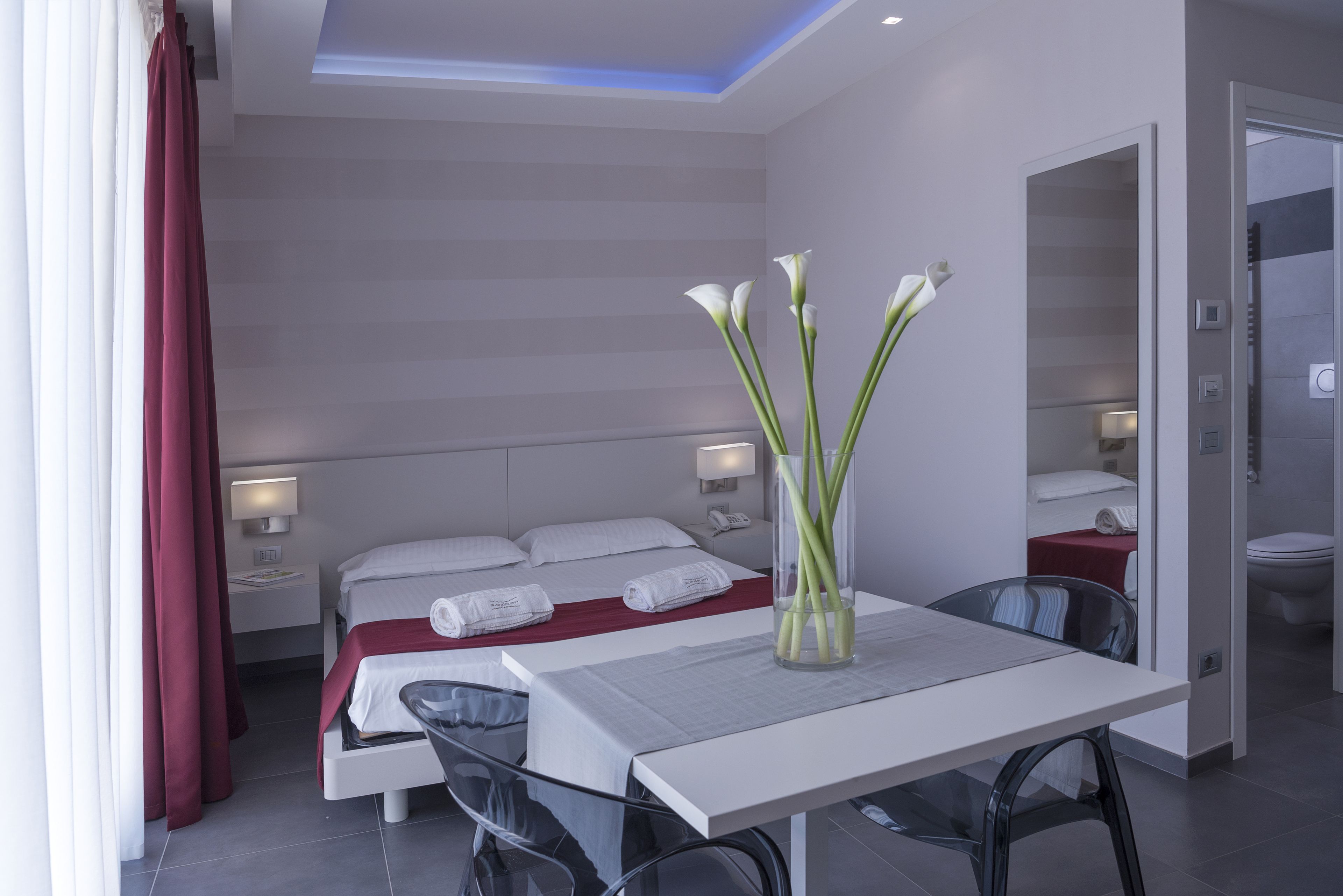 Quarto de hotel moderno com uma cama, mesas de cabeceira, uma escrivaninha com cadeiras e um vaso com flores.