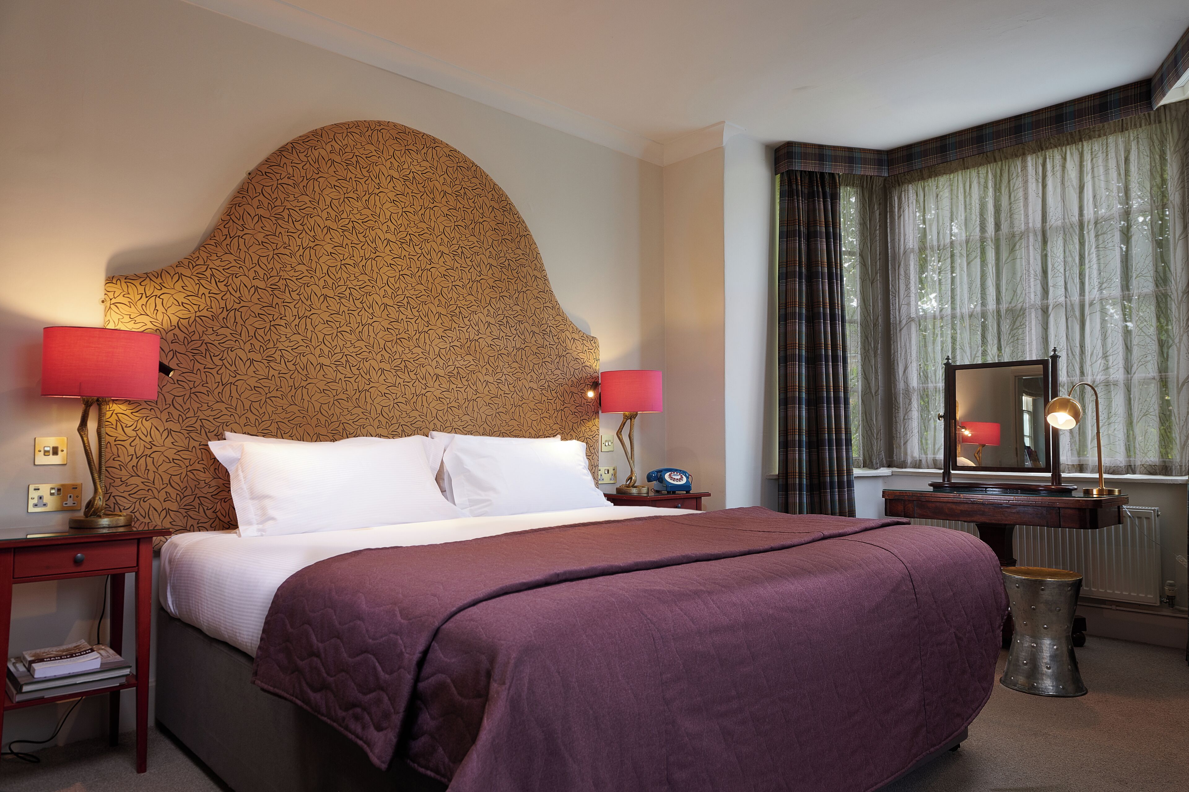 Quarto de hotel com uma cama grande, duas luminárias de cabeceira, uma mesinha de cabeceira, um espelho e uma janela com cortinas.