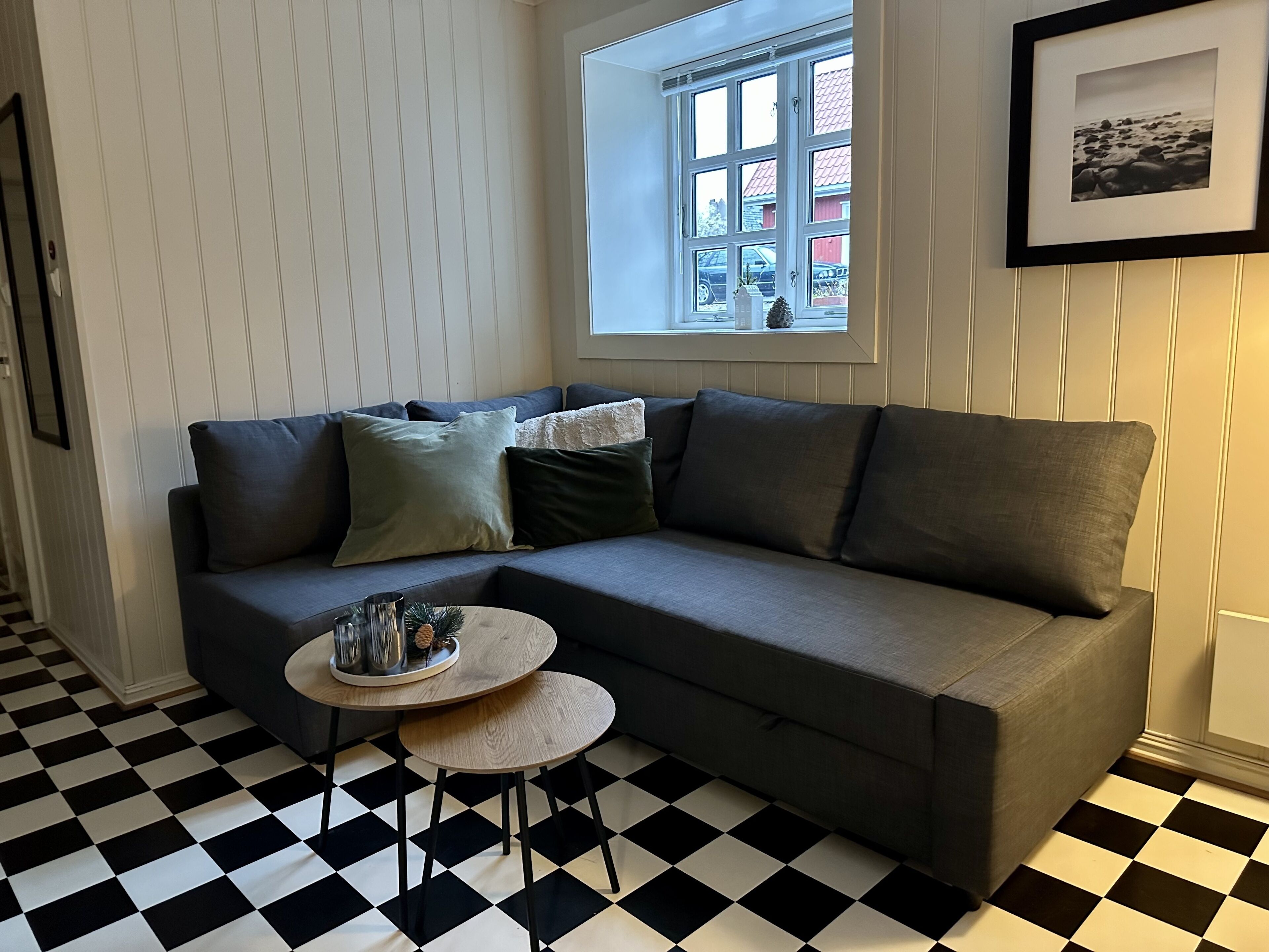 Uma sala de estar com um sofá cinzento, duas mesas redondas, um chão xadrez, uma janela com vista e um quadro na parede.