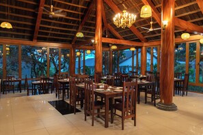 Uma sala de jantar ampla com mesas e cadeiras de madeira, janelas grandes e teto alto com vigas aparentes.