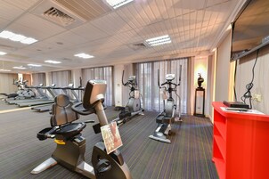 Ginásio com várias máquinas de exercício, uma receção vermelha e grandes janelas com cortinas.