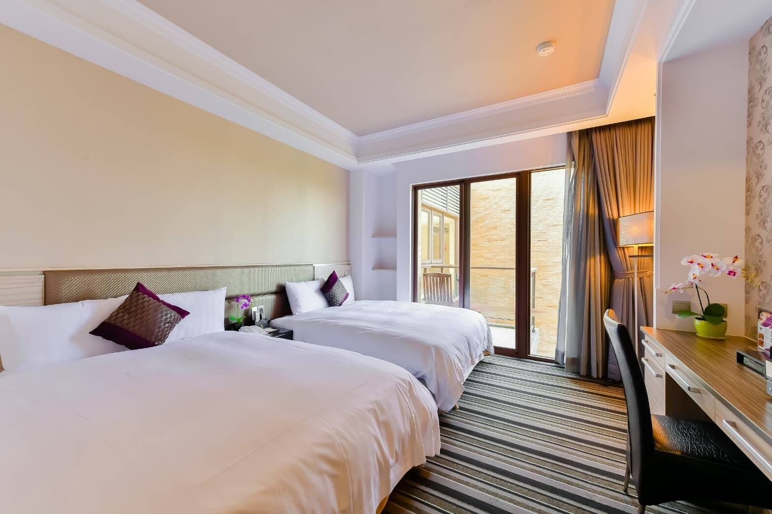 Quarto de hotel com duas camas, uma escrivaninha, uma cadeira e vista para o exterior.