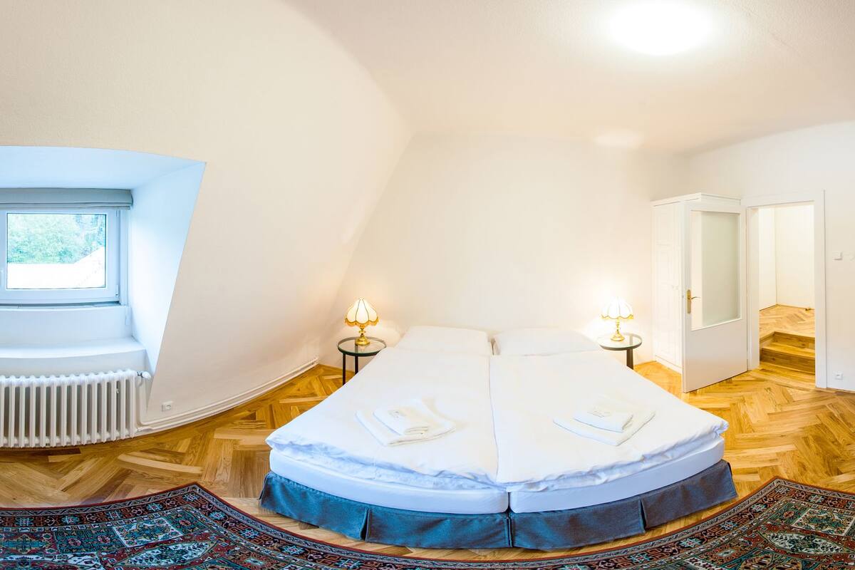 Quarto com uma janela grande, uma cama, duas mesas de cabeceira, um radiador e um vaso de plantas.