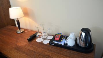 Quarto de hotel com mesa de cabeceira de madeira, comando remoto, luminária, copos de vinho, bandeja com xícaras de café e chaleira, e bandeja com sachês de chá London Gold e London Silver.