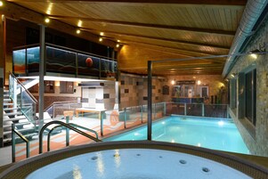 Uma piscina interior com balaustrada de vidro, uma banheira de hidromassagem e uma escadaria que leva a um mezanino.