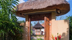 Construção com telhado de colmo e porta de madeira, cercada por vegetação e um caminho de pedra que leva a um edifício com telhado de cerâmica.
