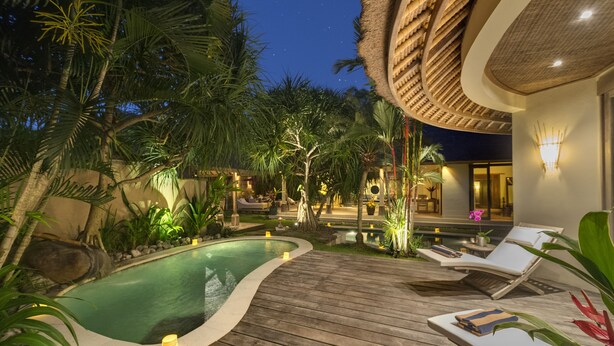 Uma área de piscina com espreguiçadeiras, uma construção com telhado de palha e um jardim bem iluminado à noite.