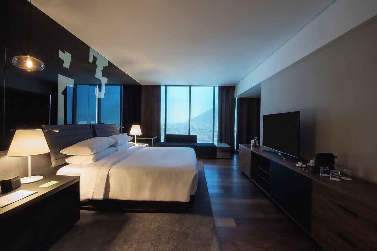 Quarto de hotel moderno com uma cama grande, abajures, televisão e vista para uma montanha.