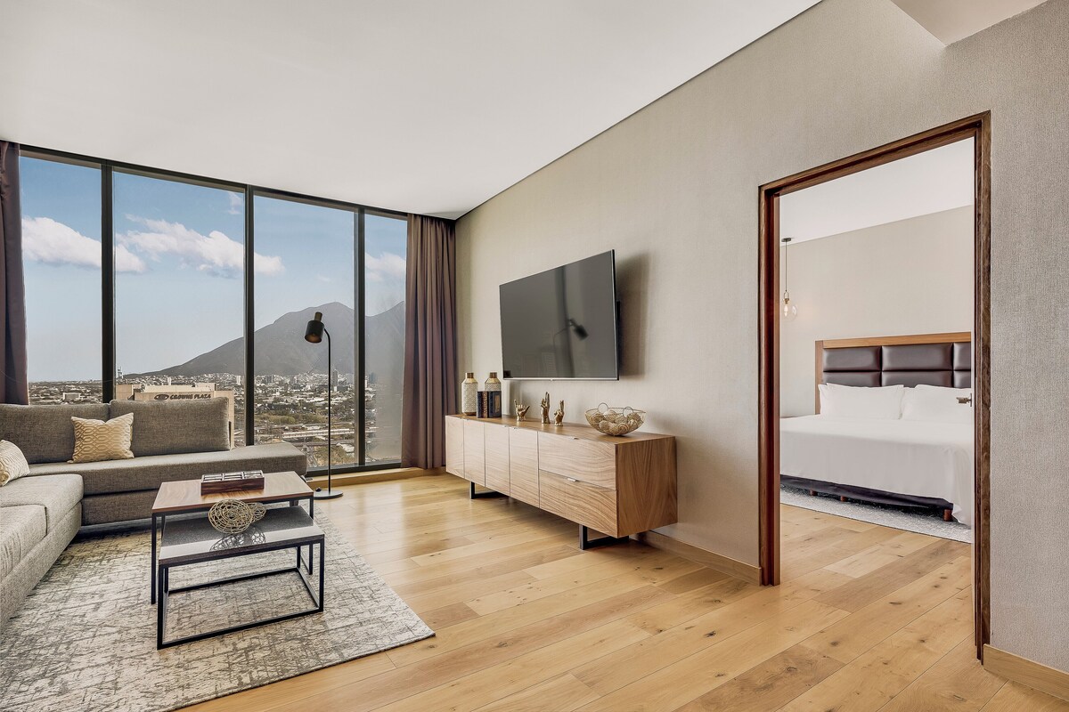 Quarto de hotel moderno com uma janela grande, TV de tela plana, sofá, mesa de centro e piso de madeira.