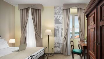 Quarto de hotel com uma cama, uma escrivaninha com um laptop, uma cadeira, um abajur e vista para a cidade pela janela.