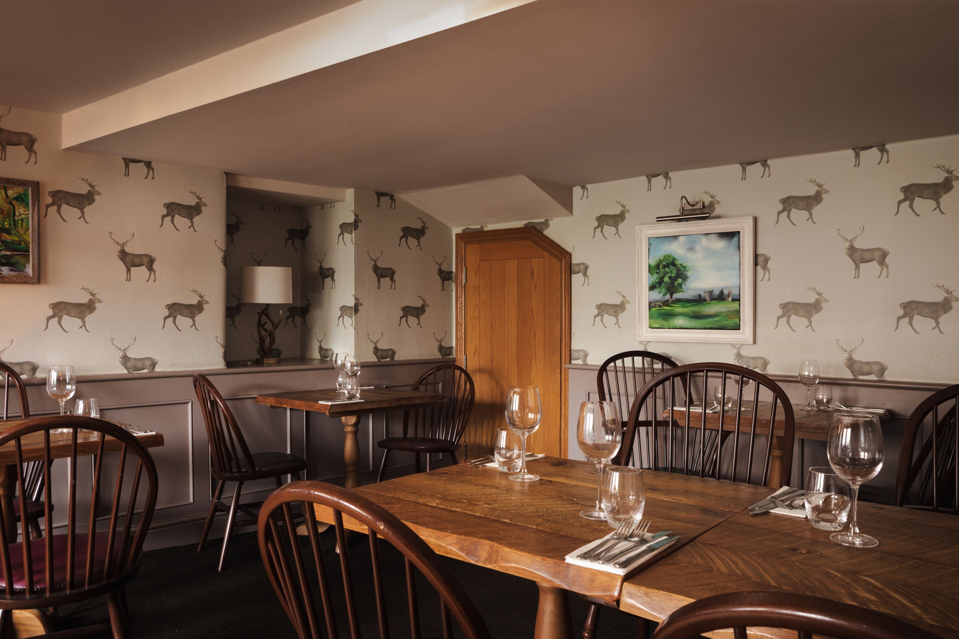 Uma sala de jantar com mesas e cadeiras de madeira, um quadro na parede e papel de parede estampado com veados.