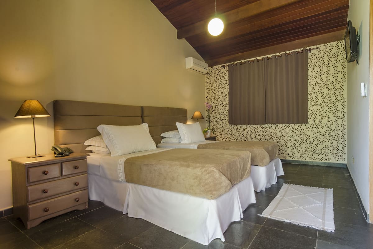 Quarto com duas camas, mesa de cabeceira, abajur e janela com cortinas.