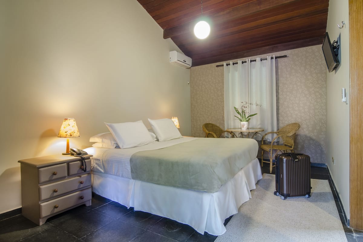 Quarto de hotel com cama, mesa de cabeceira, luminária, cadeira, mesa e mala.