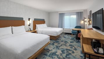 Quarto de hotel com duas camas, uma escrivaninha, uma cadeira, televisão de tela plana e uma janela grande com cortinas.