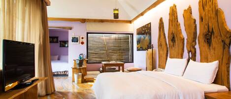 Quarto com uma cama grande, televisão sobre um suporte de madeira, janela com persianas e cabeceira de madeira.